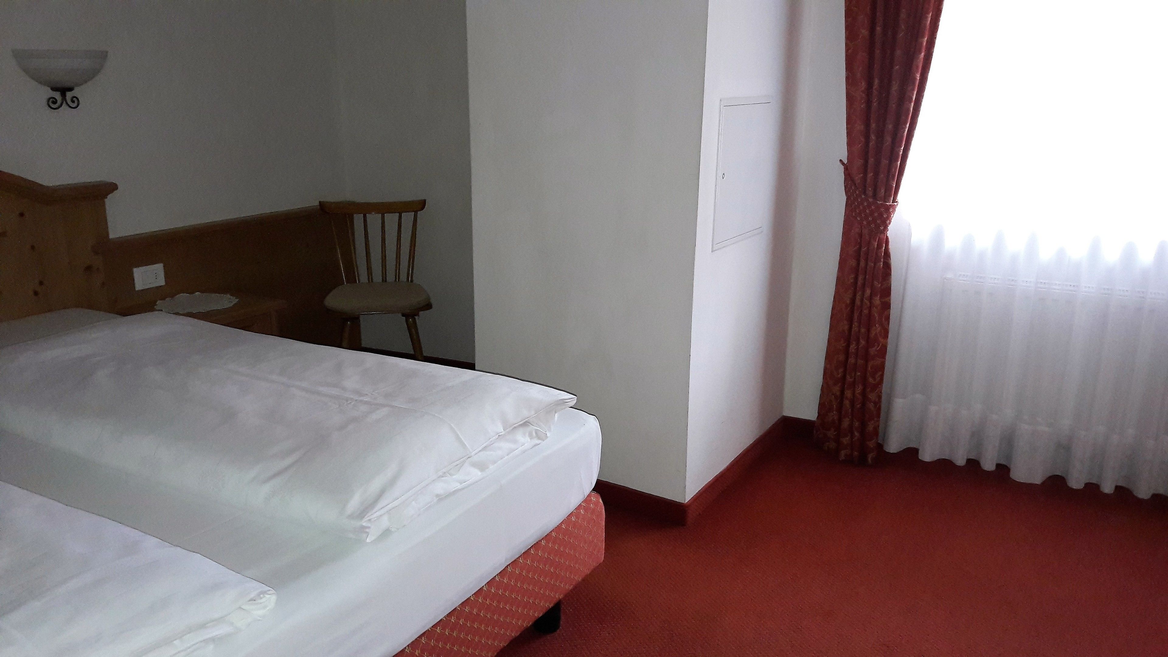 Quarto de hotel com cama, uma cadeira de madeira, luminária fixada na parede e janela com cortinas.