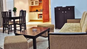 Sala de estar com sofá, um apoio para os pés e uma mesa de jantar com cadeiras. É possível ver uma área de cozinha através de uma porta aberta.