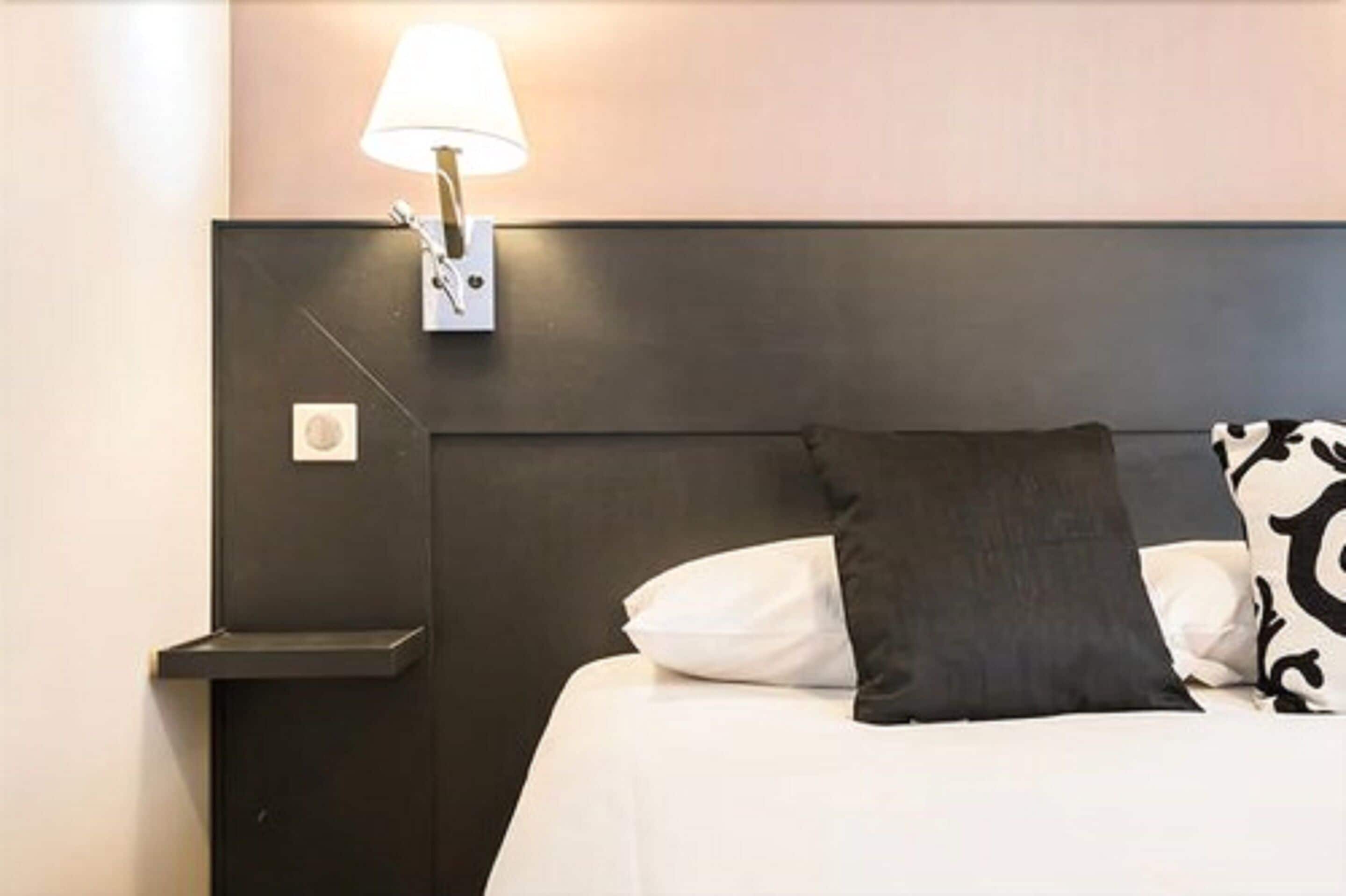 Quarto de hotel com cama, mesa de cabeceira, abajur e luminária na parede.