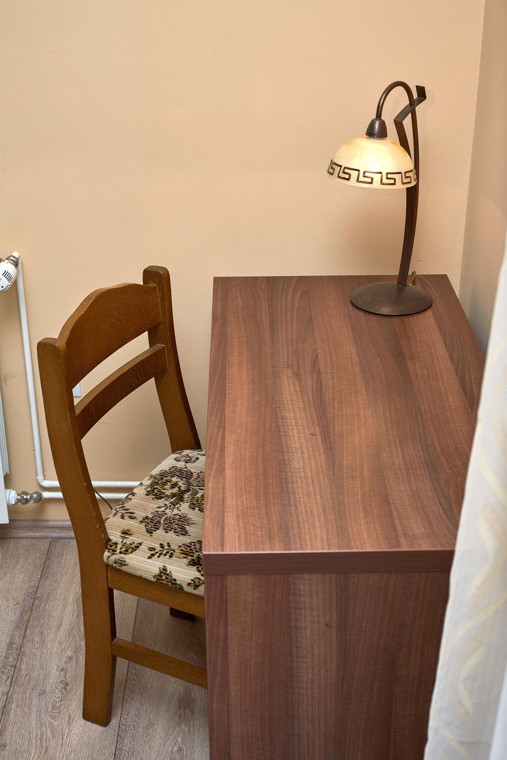 Uma mesa de madeira com uma luminária moderna, uma cadeira de madeira com almofada floral e uma parede lisa.
