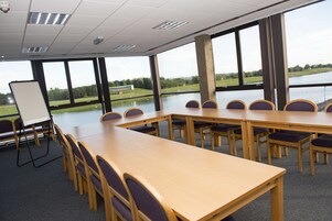 Uma sala de conferências com uma mesa grande e cadeiras, um quadro branco e amplas janelas com vista para um lago e vegetação.