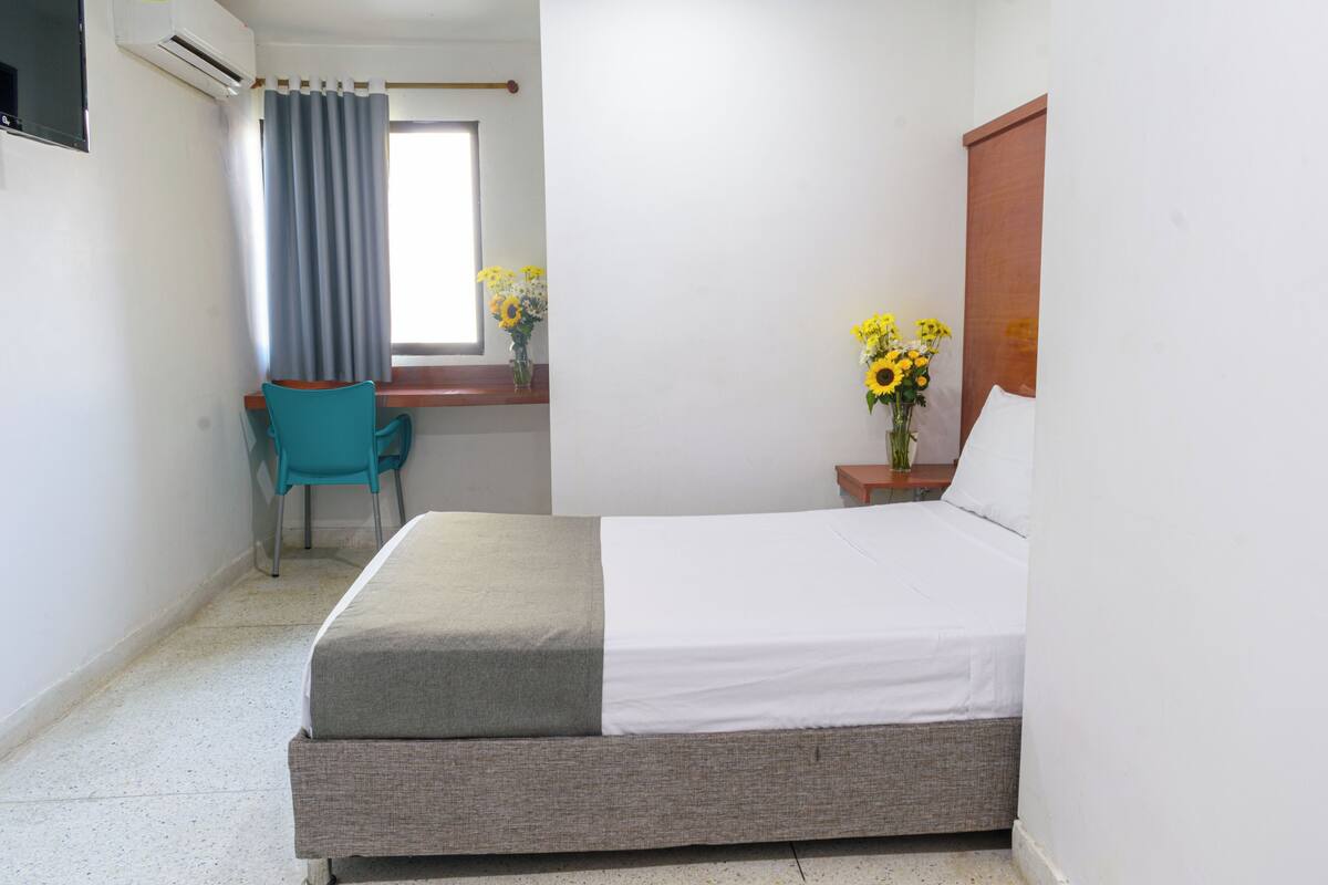 Quarto com cama, escrivaninha, cadeira e janela com cortinas.