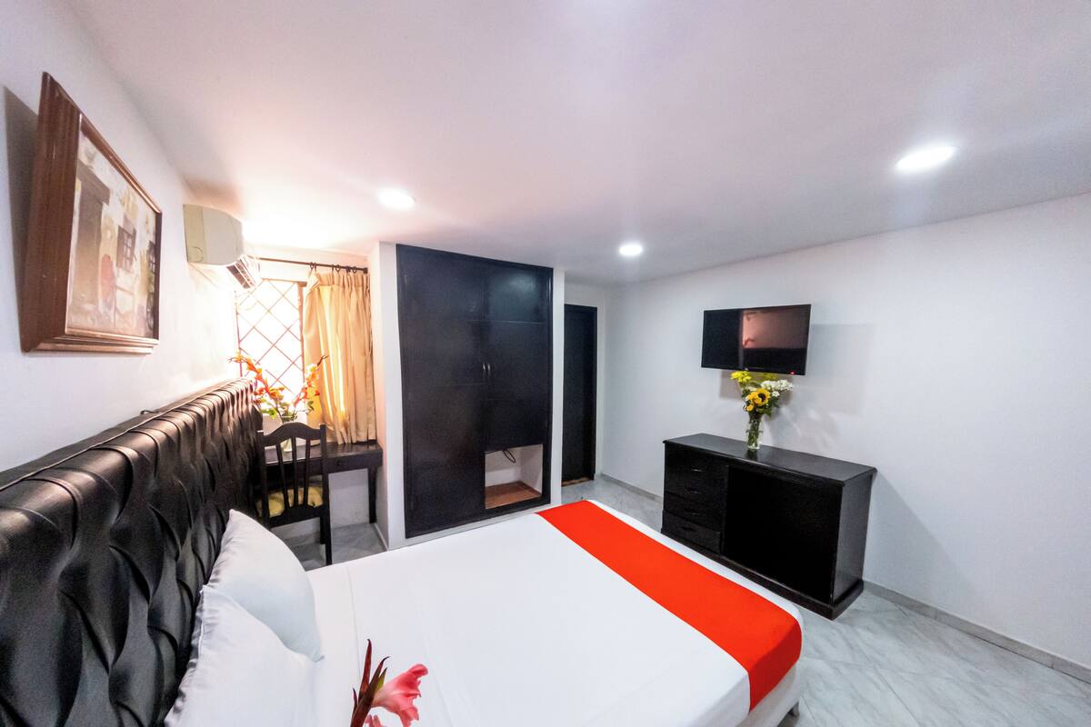 Quarto de hotel com cama, escrivaninha, guarda-roupa, televisão e janela com cortinas.
