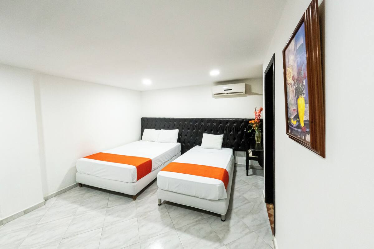 Quarto de hotel com duas camas de solteiro, cabeceira preta, ar condicionado branco, quadro emoldurado e um vaso com flores.
