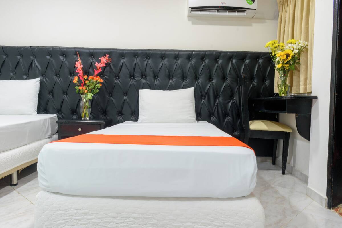Quarto de hotel com cabeceira estofada, duas camas, mesa de cabeceira com um vaso de flores, escrivaninha com cadeira e um aparelho de ar condicionado na parede.