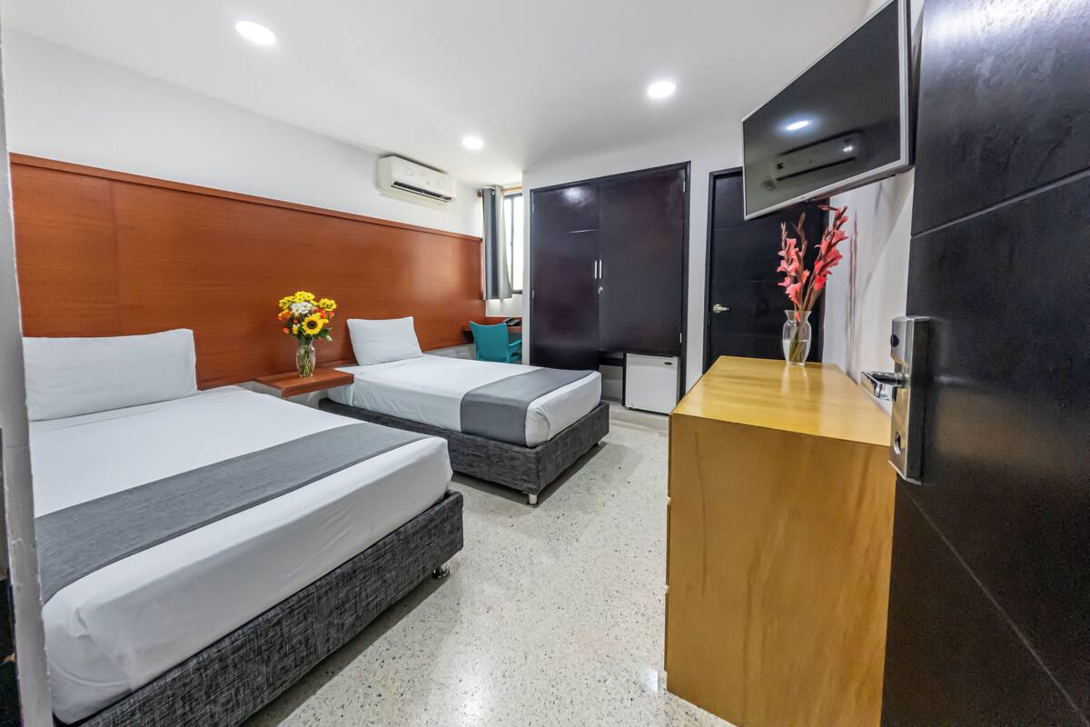Quarto de hotel com duas camas, cabeceira de madeira, uma escrivaninha pequena, televisão e guarda-roupa.