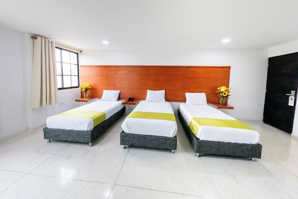 Quarto de hotel com três camas de solteiro, cabeceira de madeira, janela com cortinas e uma porta.