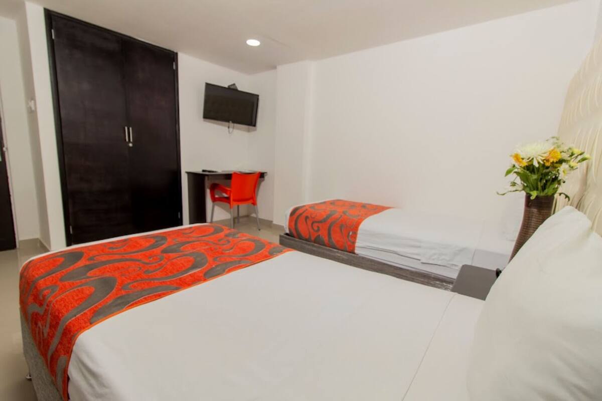 Quarto de hotel com uma cama grande, uma escrivaninha com cadeira, televisão na parede e um vaso com flores.