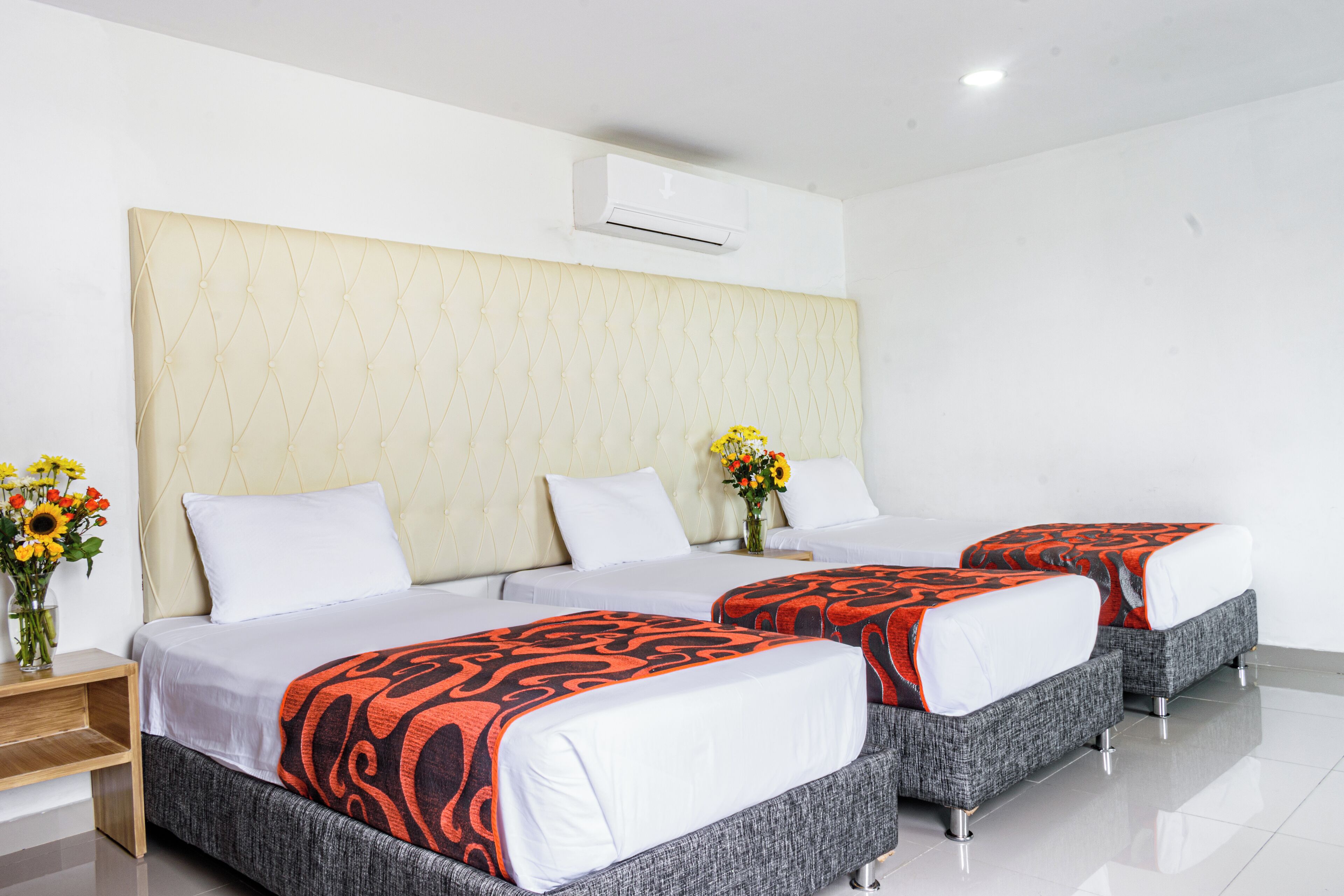 Quarto de hotel com duas camas, uma cabeceira grande, uma mesa de cabeceira com um vaso de flores e um ar condicionado.