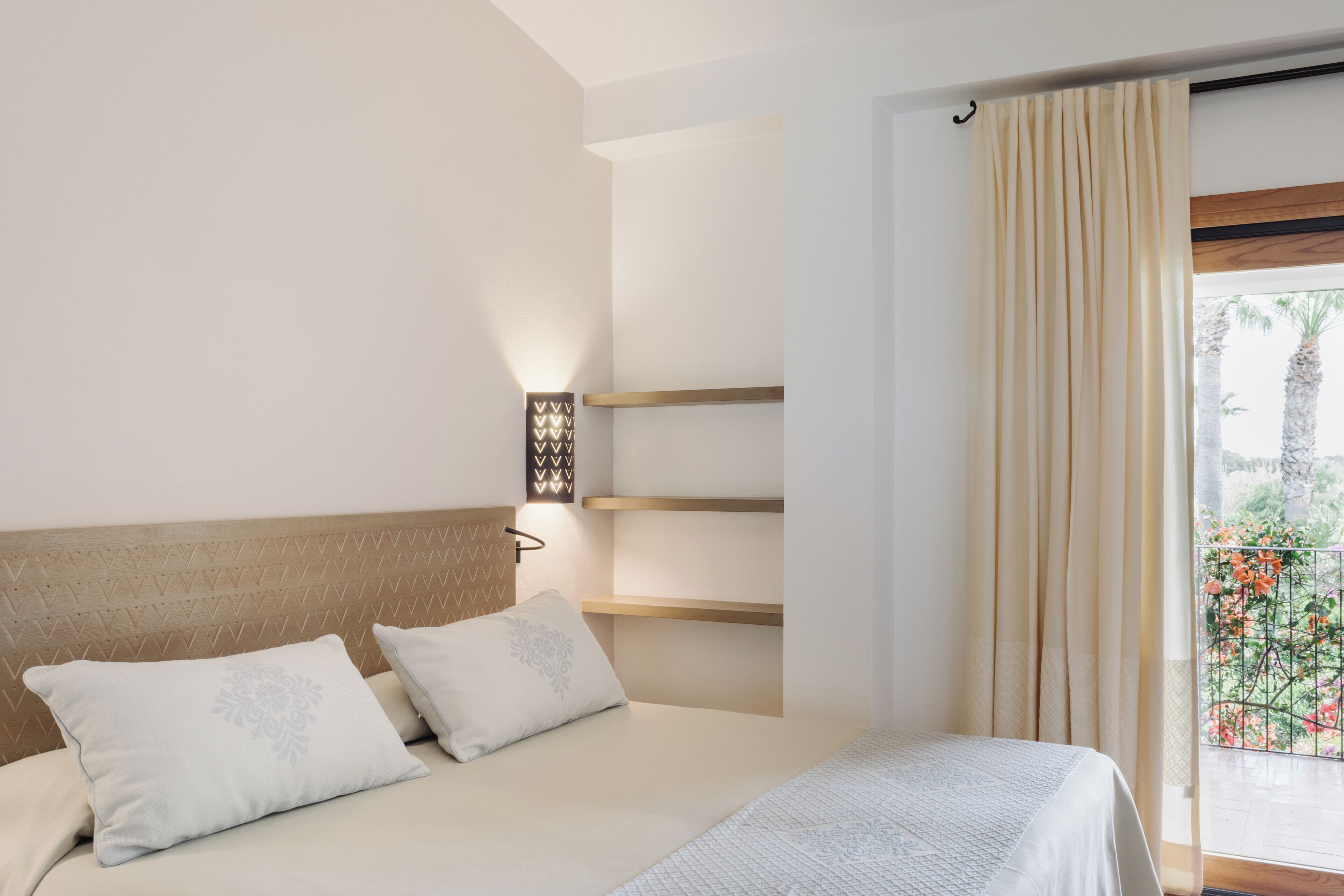 Quarto de hotel com cama, mesa de cabeceira, luminária na parede e vista para uma varanda com plantas.