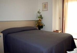 Uma cama de solteiro num quarto com parede simples, uma planta pequena e um quadro emoldurado.