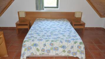 Quarto com cabeceira de madeira, duas mesas de cabeceira com luminárias, colcha florida e janela com cortinas.