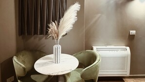 Um quarto com uma mesa redonda, duas cadeiras verdes, um vaso com plumas de capim-do-campo, uma cortina e um aparelho de ar condicionado branco.