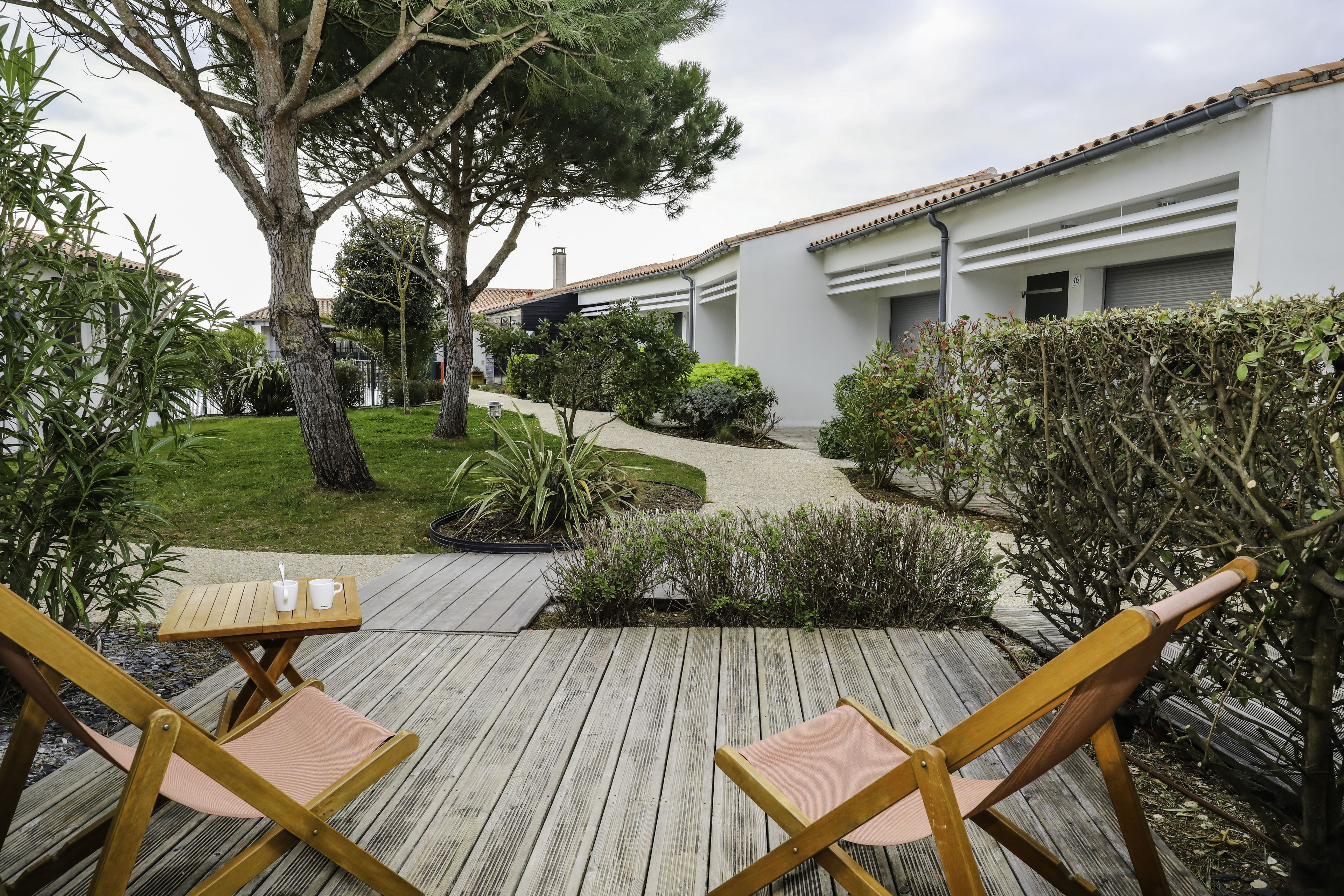 Um deck de madeira com duas cadeiras de madeira e uma mesa, cercado por um jardim com diversas plantas e um caminho que leva a um edifício com telhado de azulejos.