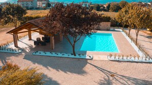Uma piscina com área de estar coberta, cercada por árvores e vista para edifícios ao longe.