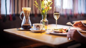 Uma mesa com um coador de café, uma xícara, um copo de vinho branco, um vaso com flores e um prato com comida.