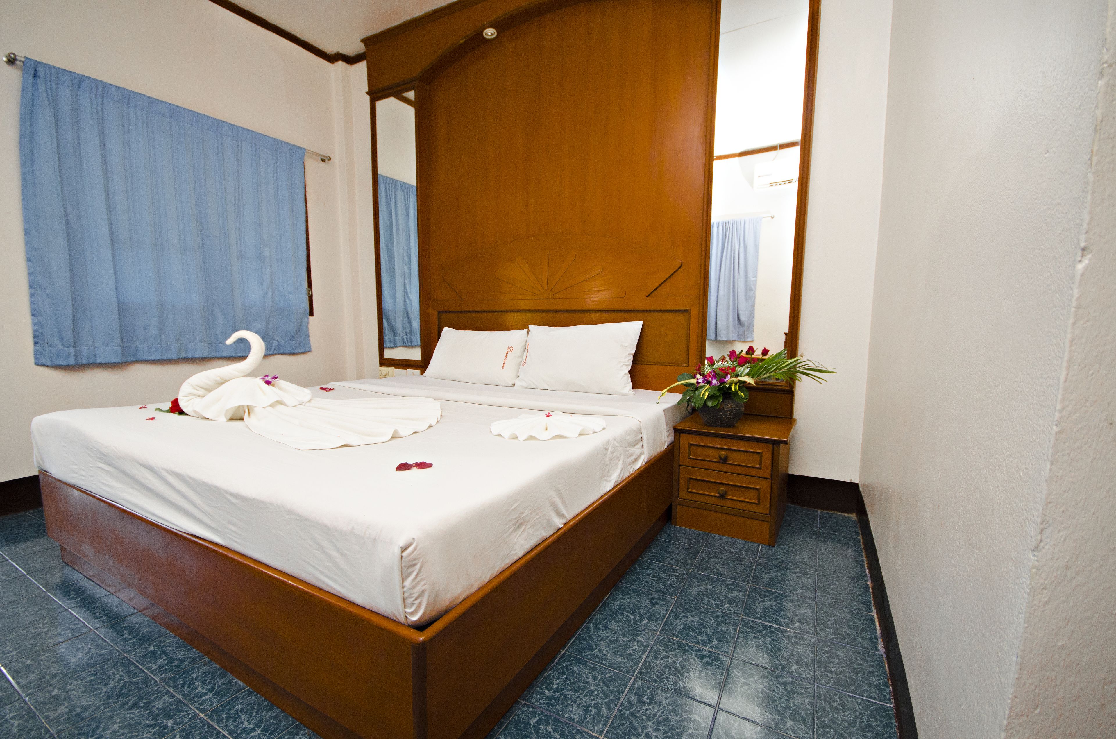 Cama bem arrumada com lençóis brancos e almofadas decorativas em formato de cisne, cabeceira de madeira, mesa de cabeceira com planta em vaso e um espelho grande na parede.