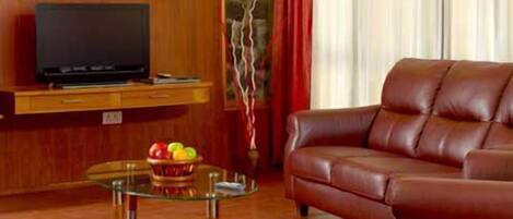 Um sofá de couro marrom, uma mesa de centro de vidro com frutas e uma televisão sobre um suporte de madeira.