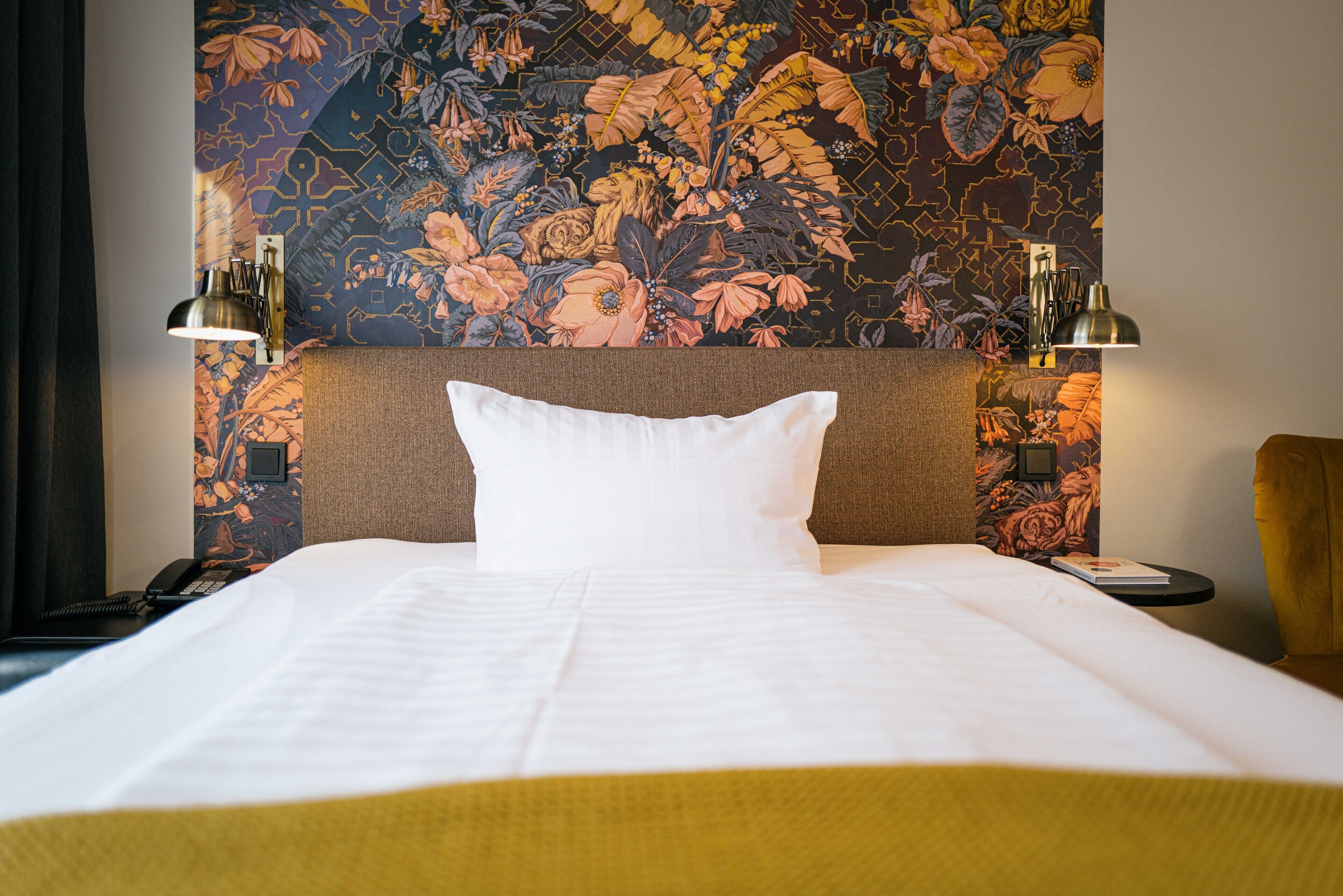 Uma cama bem arrumada com lençóis brancos, cabeceira com padrão floral e duas luminárias de cabeceira.