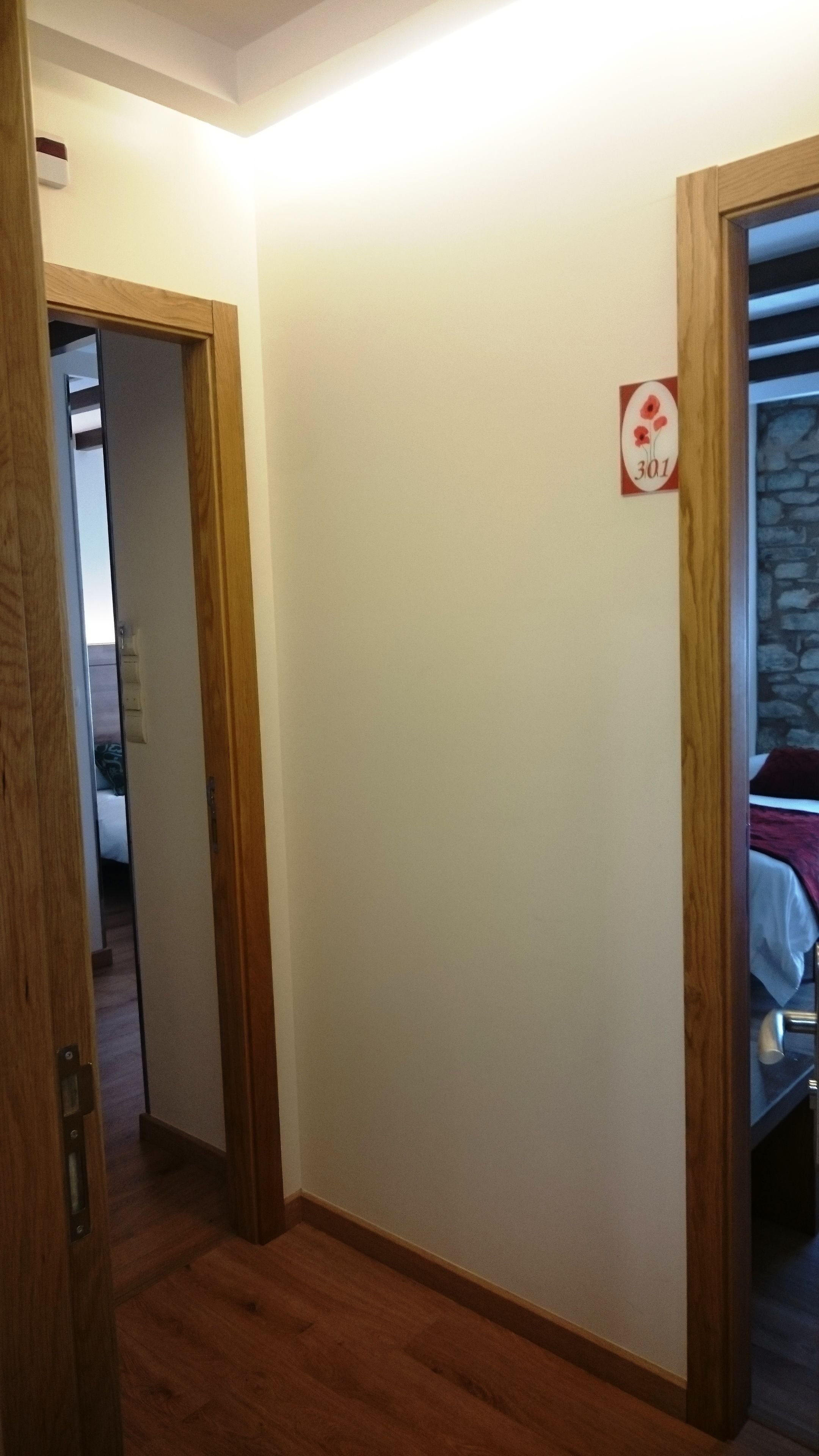 Um corredor com piso de madeira e uma porta que dá para um quarto com parede de pedra. Uma placa com o número 301 é visível na parede.