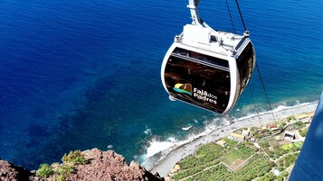 Um teleférico com um logotipo e o texto "Fajões dos Padres" sobre uma paisagem costeira.