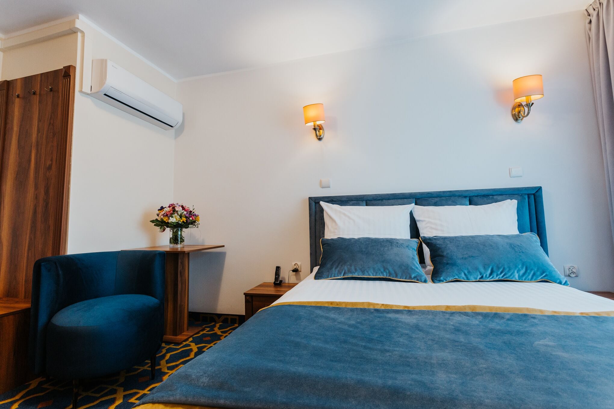 Quarto de hotel com cama, cadeira, ar condicionado, mesa de cabeceira e duas luminárias fixadas na parede.