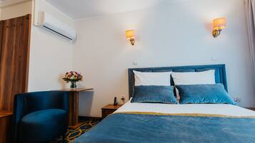 Quarto de hotel com cama, cadeira, ar condicionado, mesa de cabeceira e duas luminárias fixadas na parede.