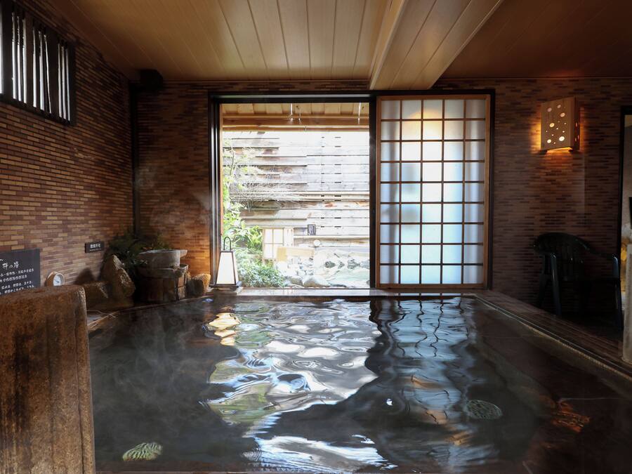 Um onsen japonês tradicional com interior de madeira, uma bacia de pedra e vista para o exterior através de uma porta deslizante.