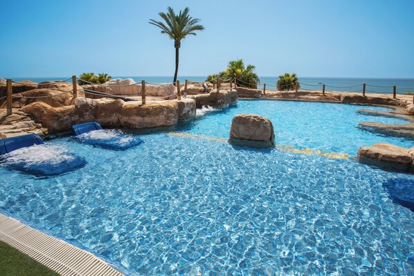 Piscina azul límpida com formações rochosas e palmeiras, cercada por uma cerca de madeira.