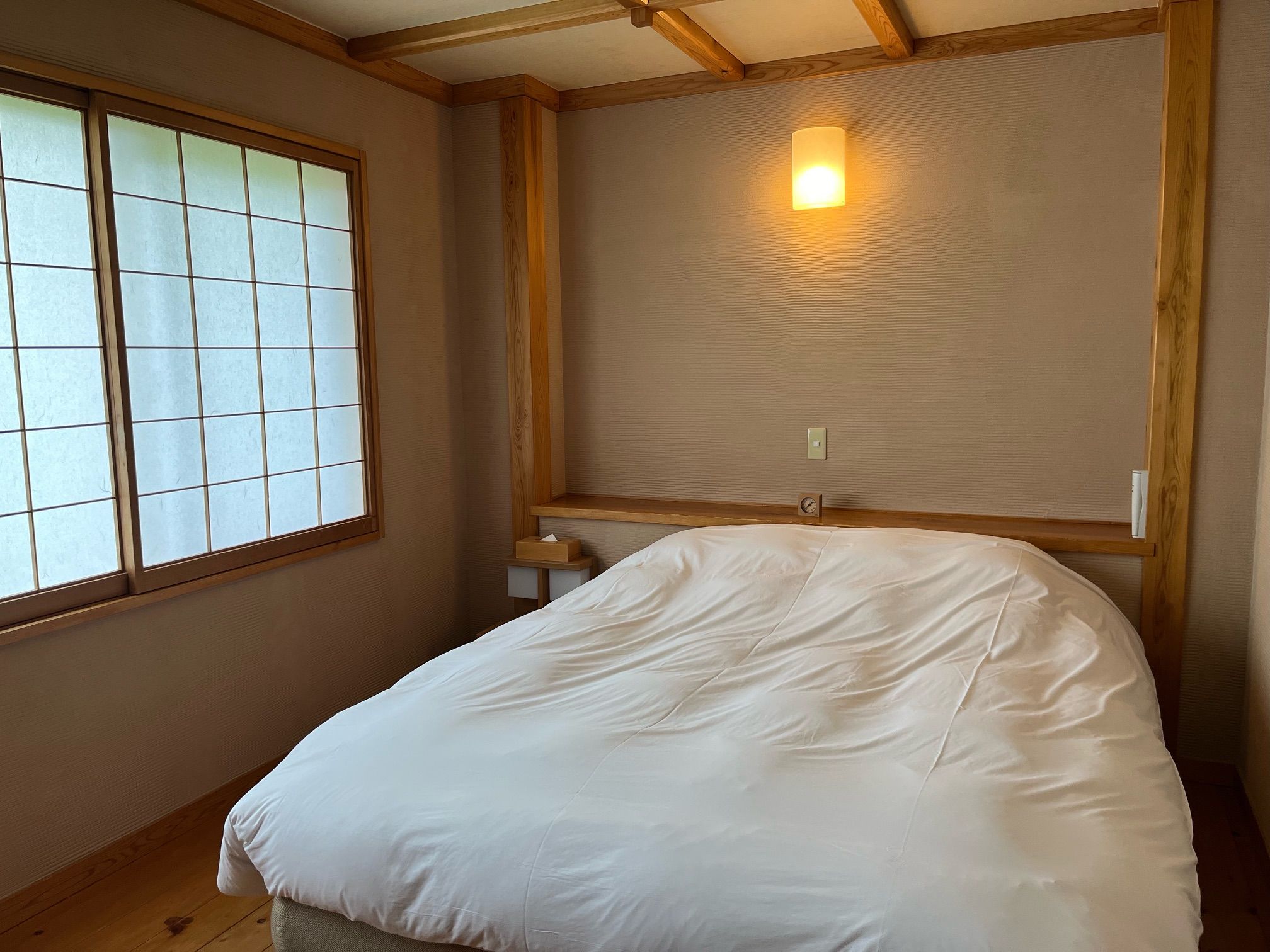 Quarto com cama, janela de vidro fosco, vigas de madeira no teto e luminária fixada na parede.