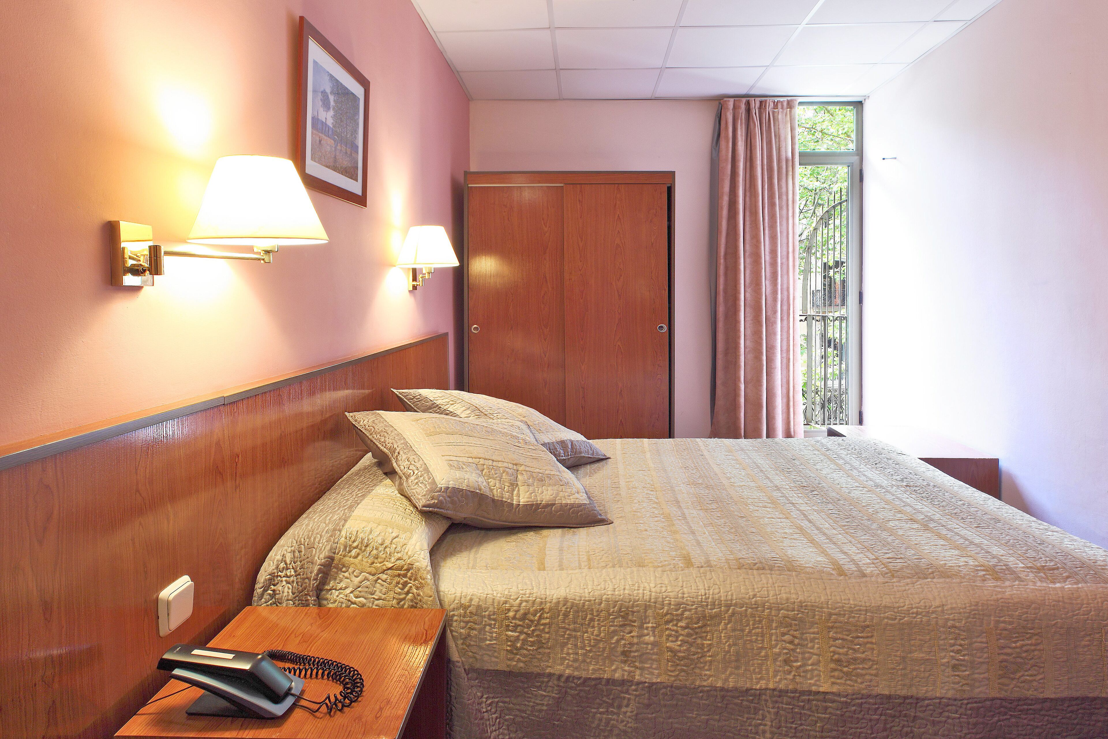 Quarto de hotel com cama, duas luminárias de cabeceira, telefone e janela com cortinas.