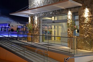 Entrada moderna de hotel com fachada de pedra, portas de vidro e área de estar ao ar livre.