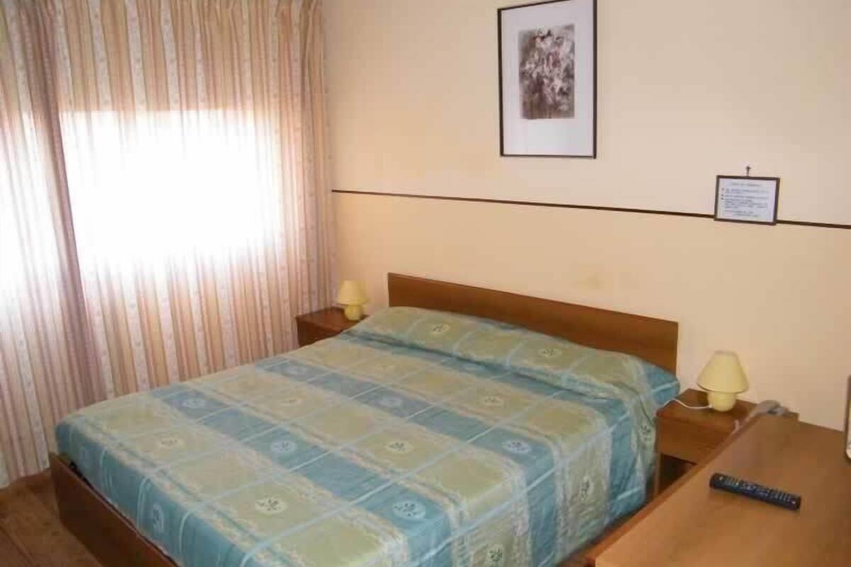 Quarto de hotel com cama, mesa de cabeceira, luminária, quadro na parede e janela com cortinas.