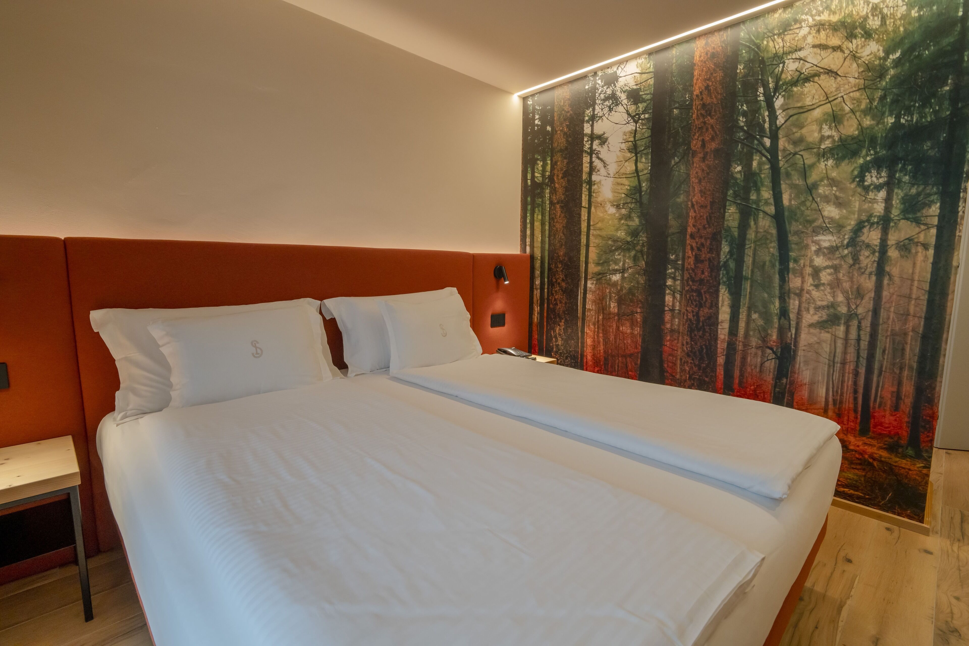 Quarto de hotel moderno com uma cama grande, cabeceira laranja e um mural de parede com tema de floresta.