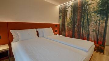Quarto de hotel moderno com uma cama grande, cabeceira laranja e um mural de parede com tema de floresta.