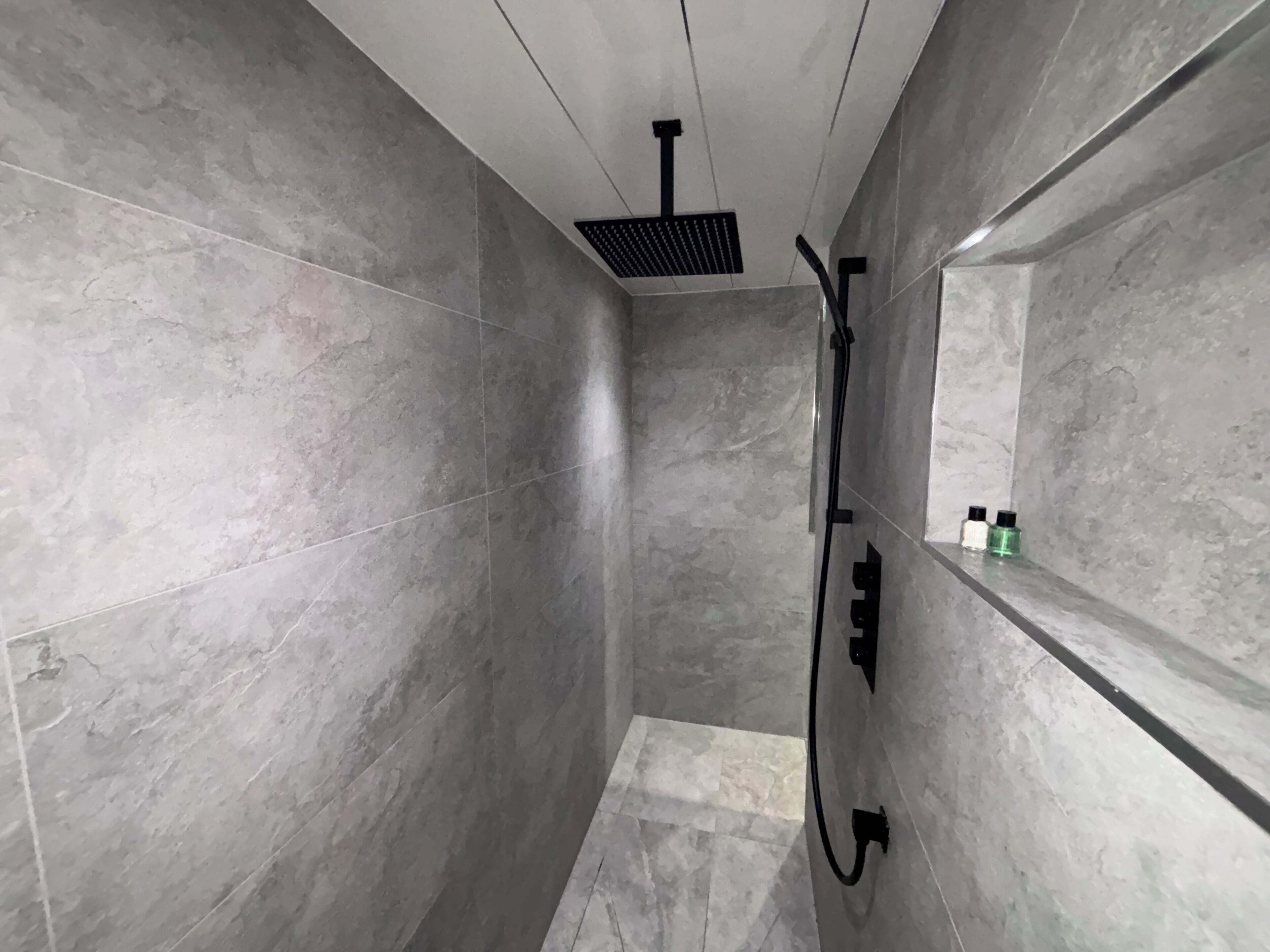 Um espaço de banho moderno com azulejos cinzentos, um chuveiro preto e um pequeno nicho para produtos de higiene.