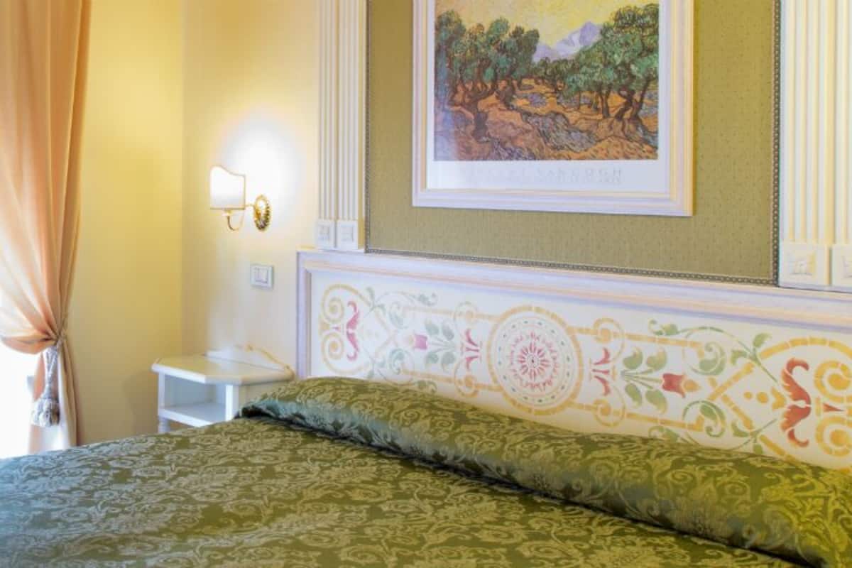 Quarto de hotel com cama, cortinas, mesa de cabeceira, luminária na parede e um quadro emoldurado.