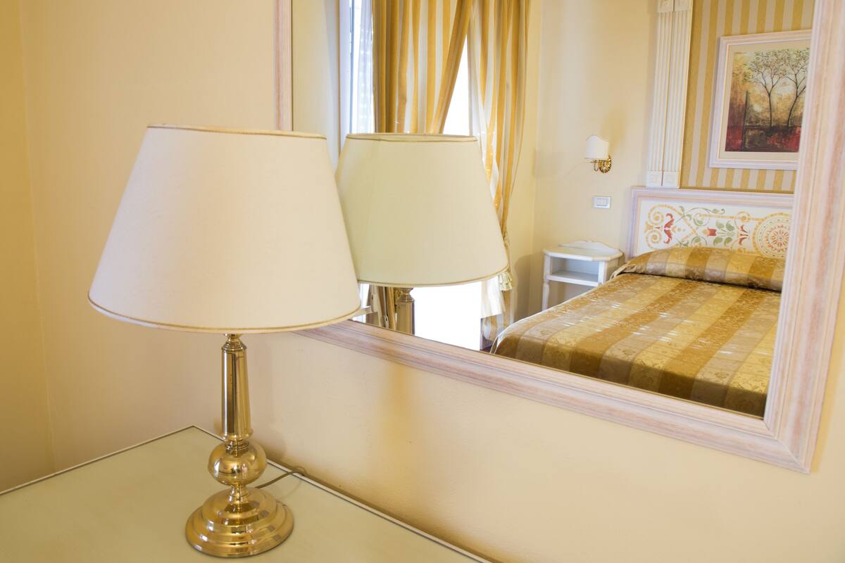 Quarto de hotel com cama, duas luminárias de cabeceira, mesa de apoio e luminária fixada na parede.