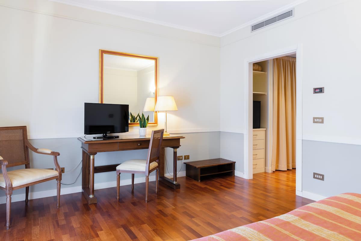 Quarto de hotel com escrivaninha de madeira, televisão, cadeira, cama, espelho, abajur e armário.