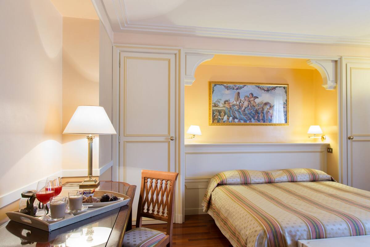 Quarto de hotel com uma cama, uma mesa com bandeja de itens de café da manhã, uma cadeira e um quadro na parede.