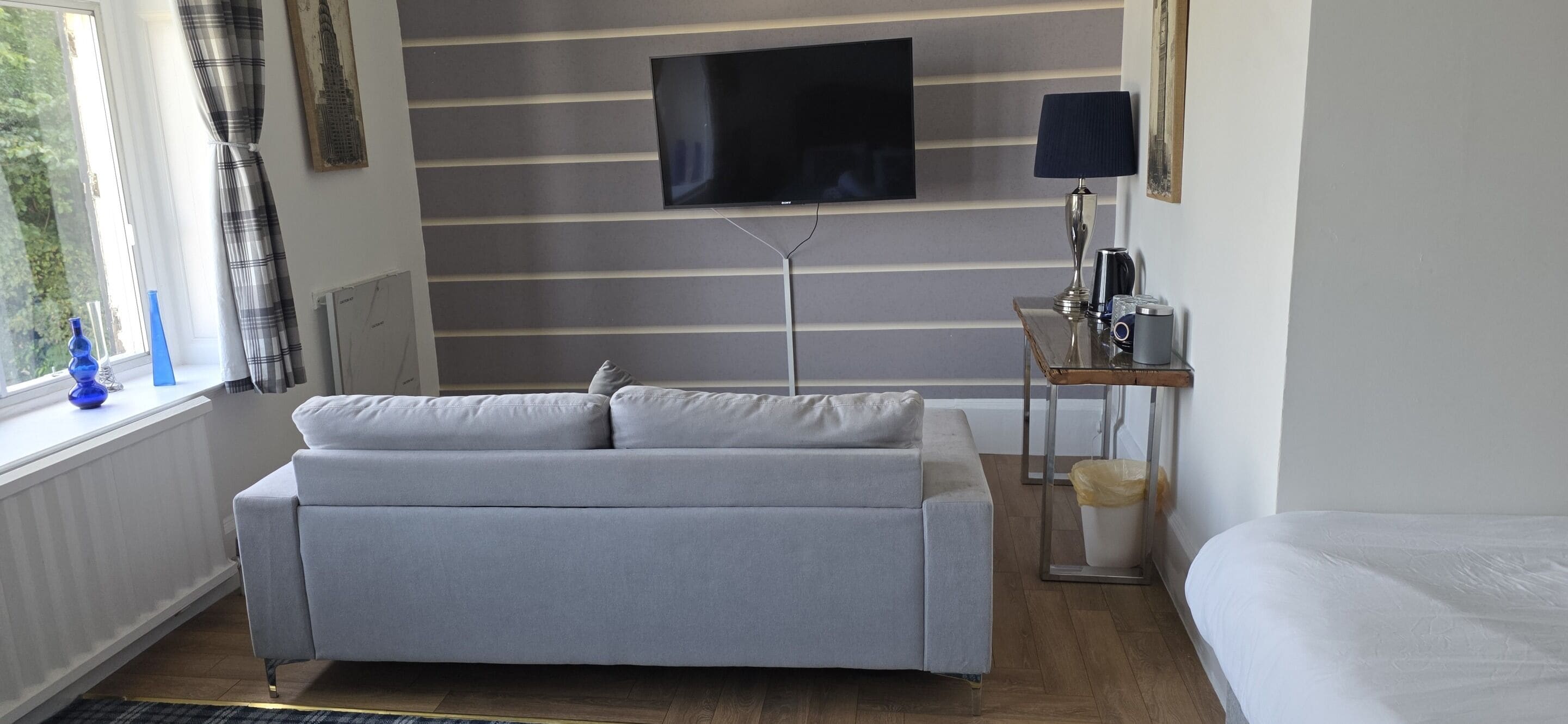 Quarto de hotel moderno com sofá cinza, televisão de tela plana, mesa de cabeceira com abajur e janela com cortinas.