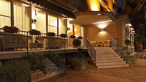 Um hotel com fachada de tijolos, área externa com mesas e cadeiras, e escadaria que leva à entrada.