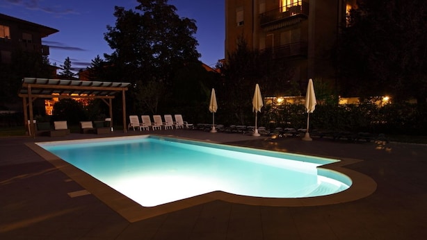 Piscina com espreguiçadeiras e guarda-sóis, cercada por edifícios e árvores.