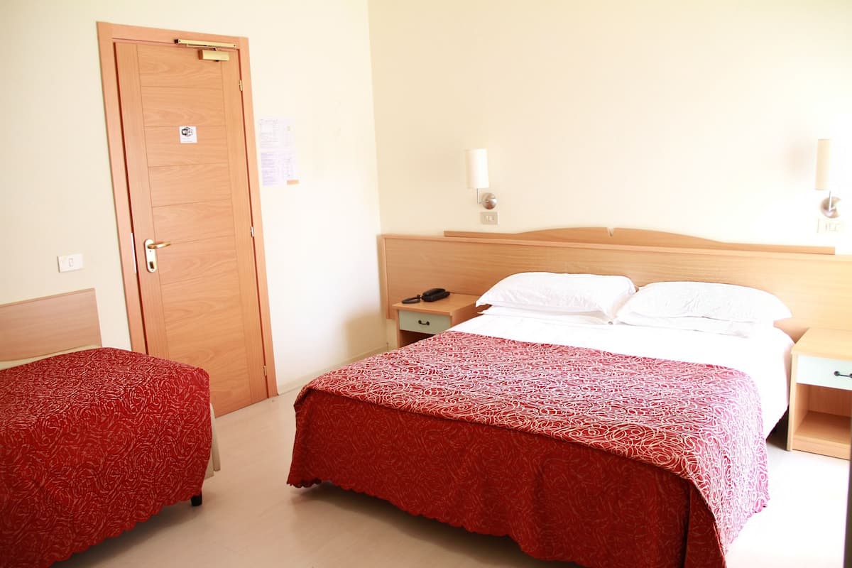 Quarto de hotel com porta de madeira, cama com colcha estampada vermelha, mesa de cabeceira com abajur e criado-mudo com telefone.