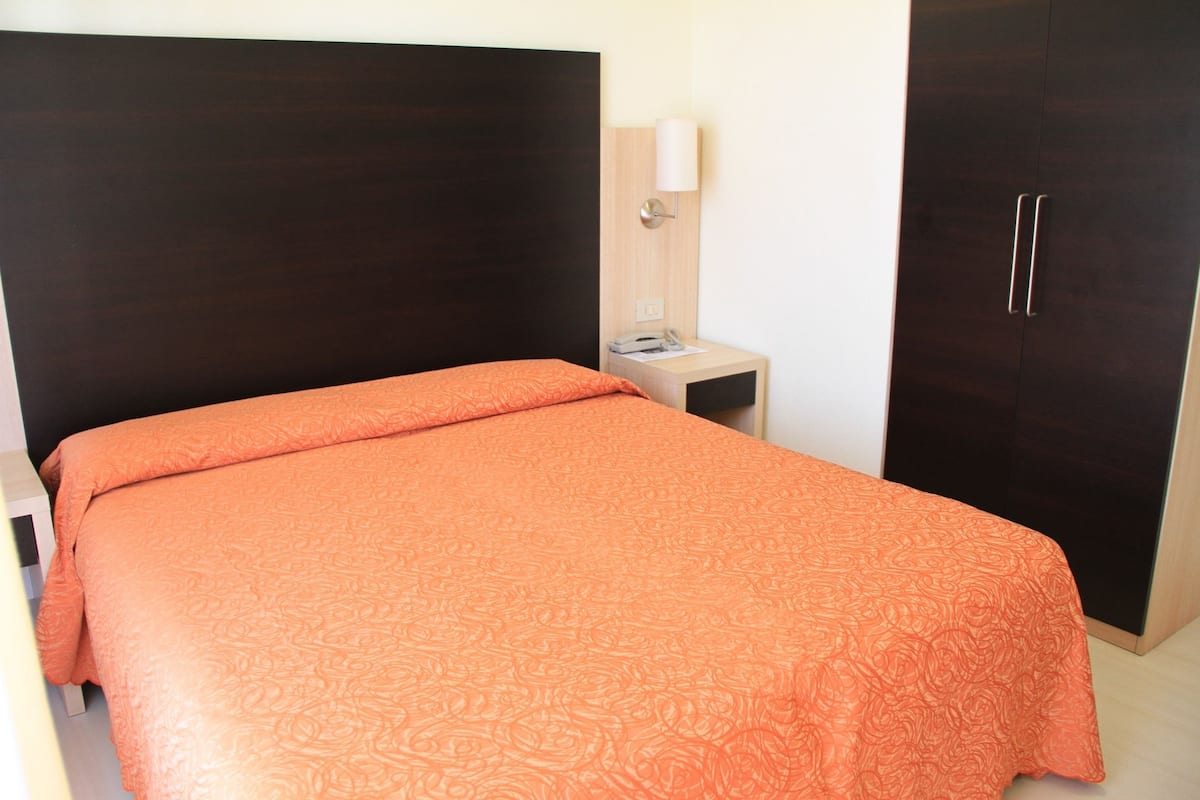 Cama bem arrumada com colcha laranja estampada, cabeceira de madeira escura, mesa de cabeceira com abajur e guarda-roupa com duas portas.