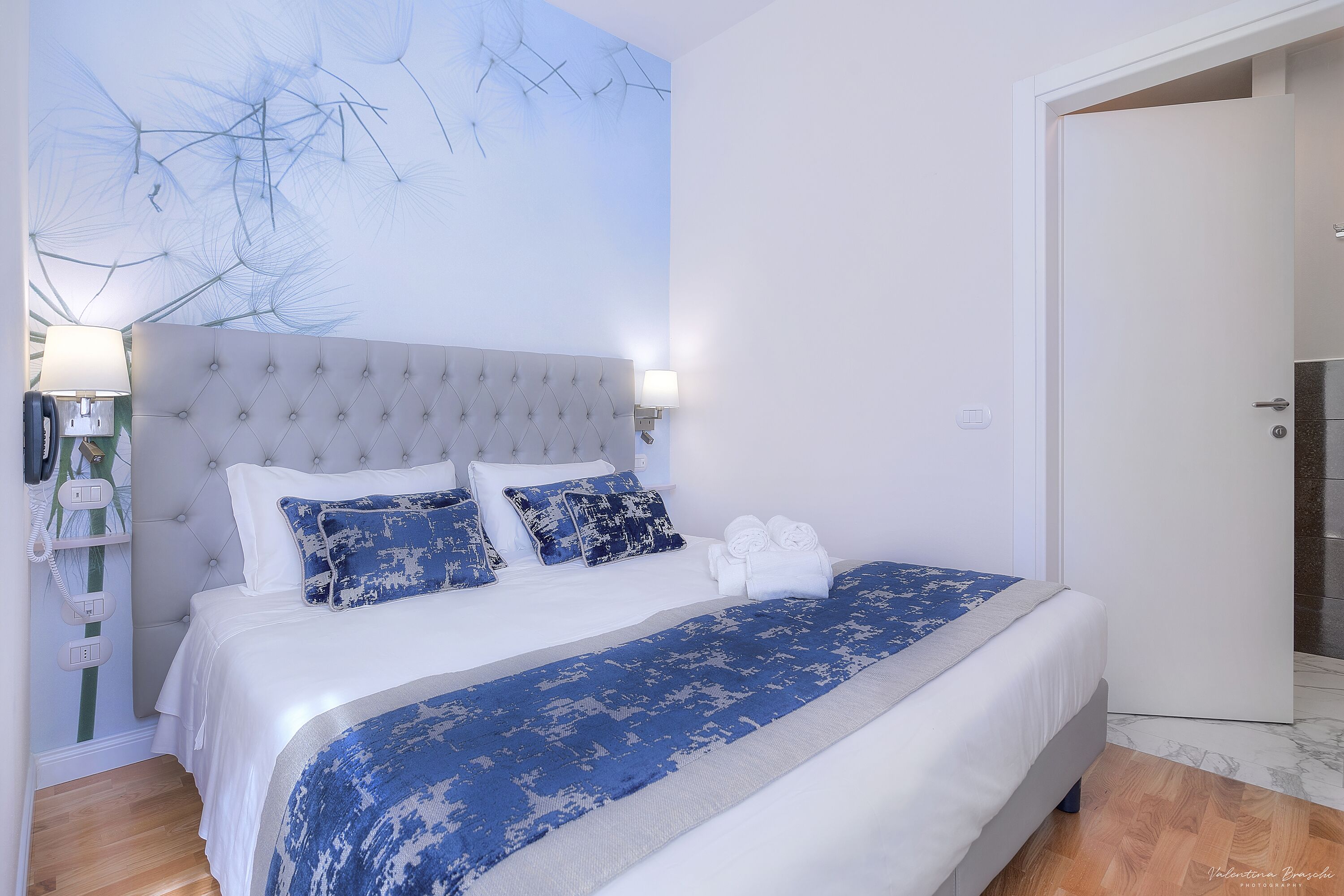 Quarto com uma cama grande, luminária na parede e porta que dá para outro cômodo.