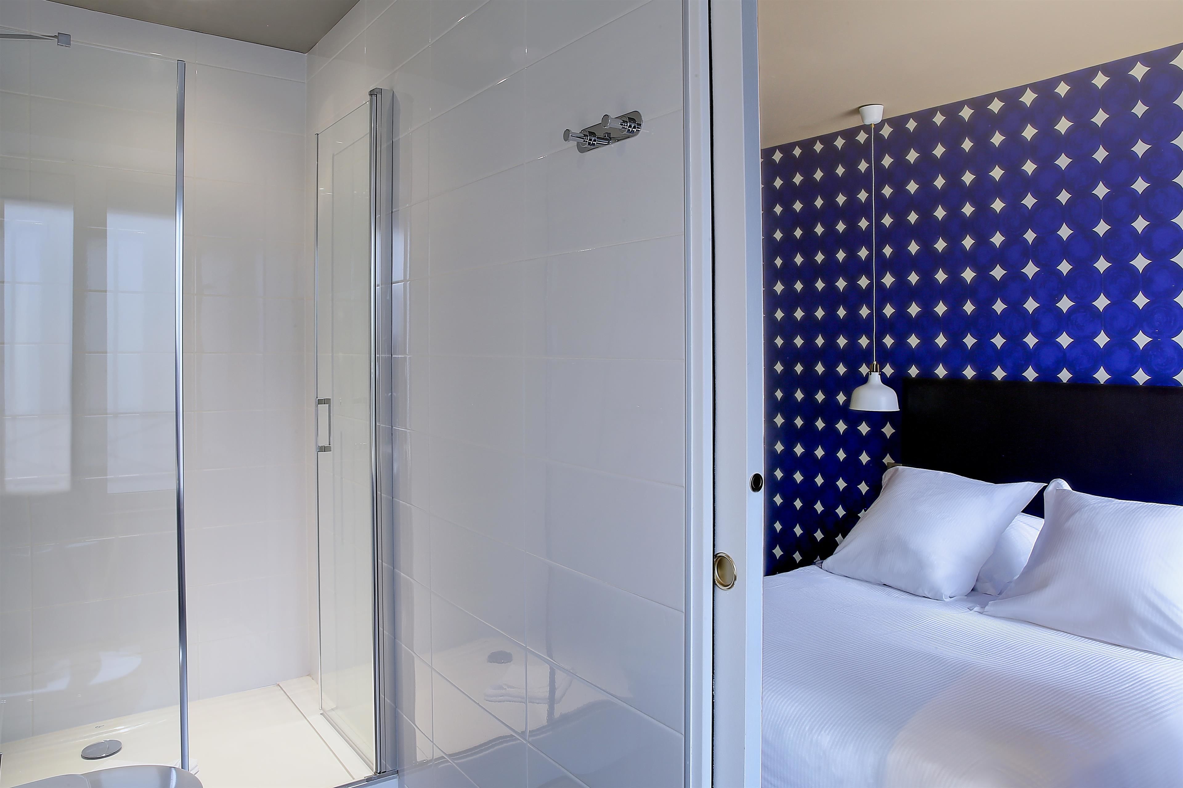 Um banheiro moderno com box de vidro e cama com roupa de cama branca e cabeceira estampada em azul.