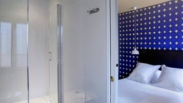 Um banheiro moderno com box de vidro e cama com roupa de cama branca e cabeceira estampada em azul.