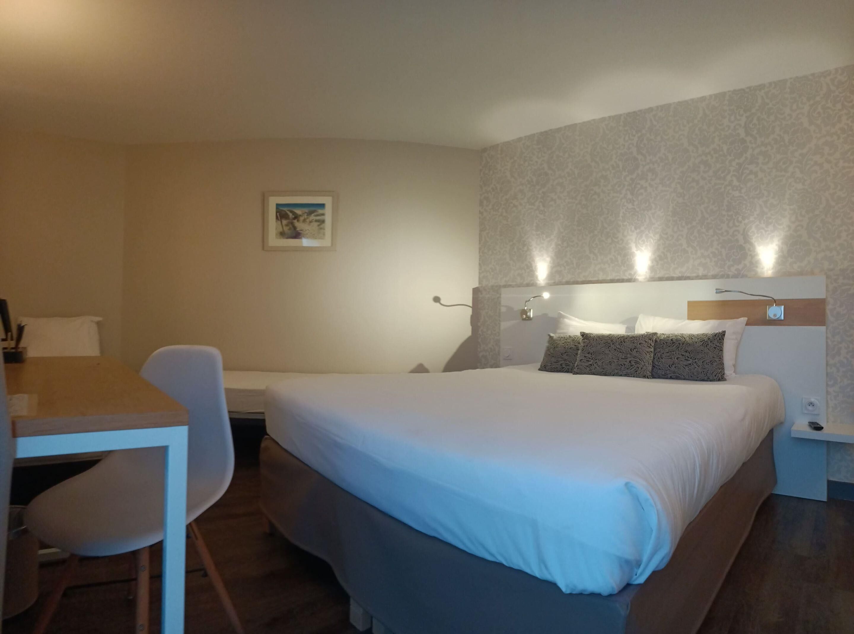 Quarto de hotel com cama, mesa de cabeceira, luminária e escrivaninha com cadeira.