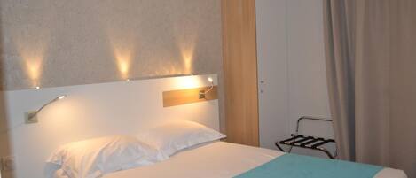 Quarto de hotel com cama, mesa de cabeceira, luminária na parede, porta-toalhas e cortina.